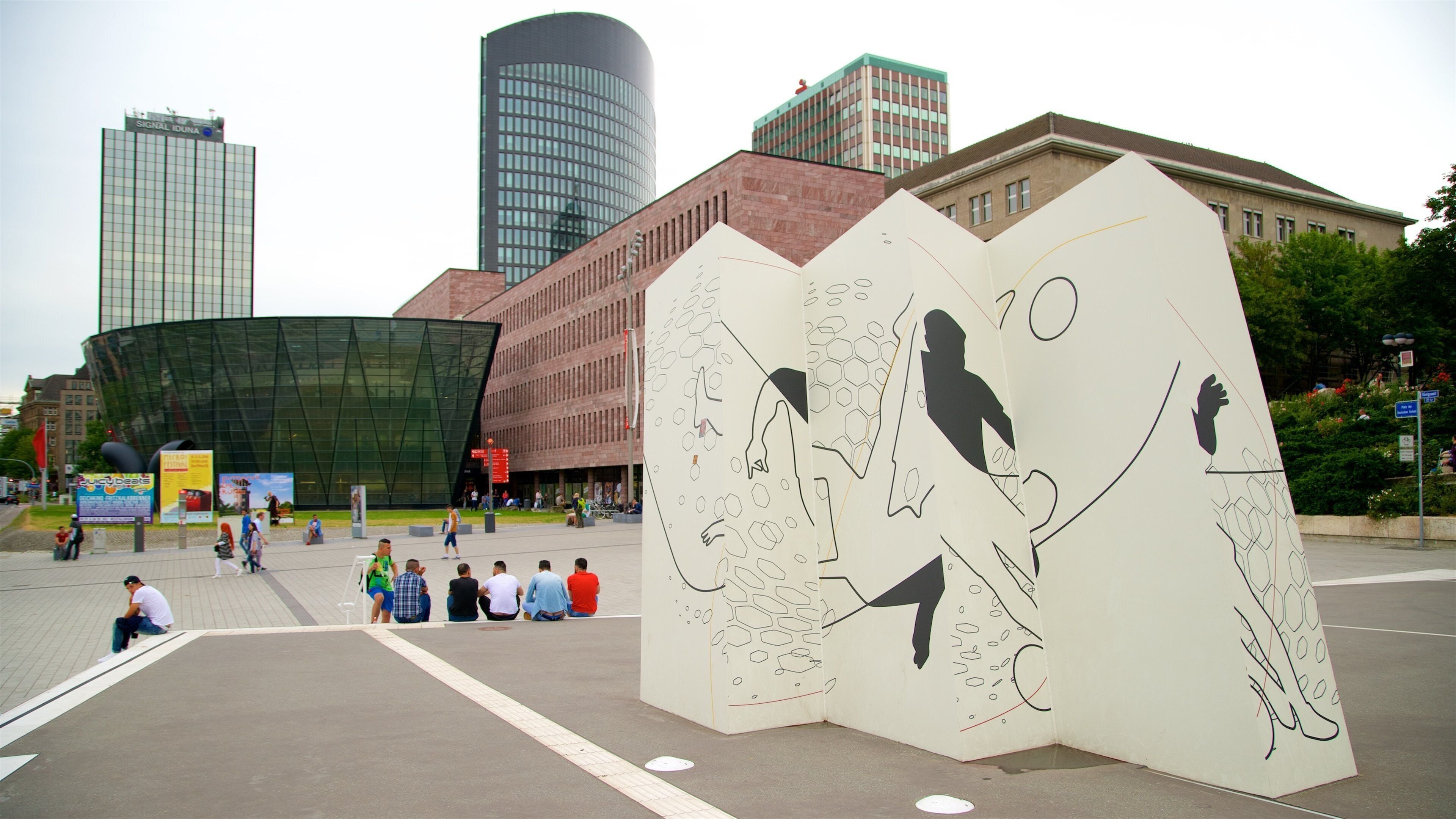 Uma praça urbana moderna com um grande edifício de vidro, um edifício de tijolos vermelhos e uma escultura de figuras humanas abstratas.