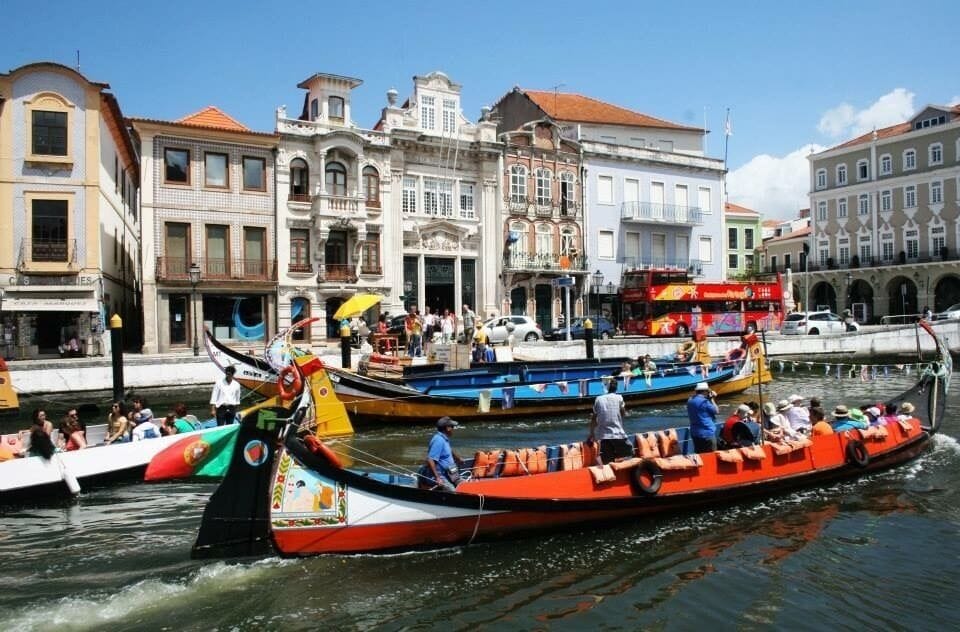 Um desfile de barcos tradicionais em um rio, com espectadores e edifícios ricamente decorados ao fundo.