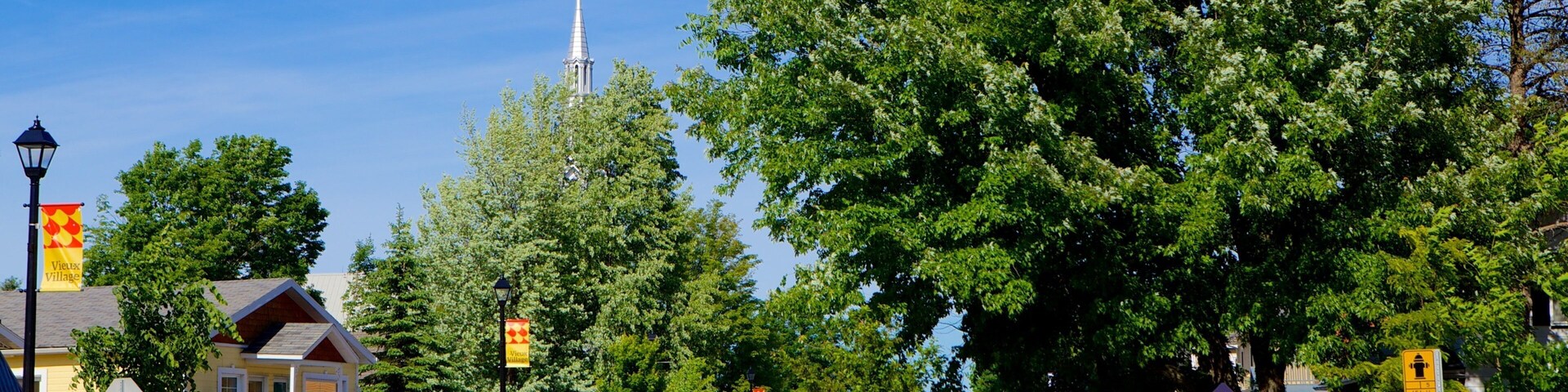 Rua arborizada com um campanário de igreja e um pequeno edifício com placa amarela.