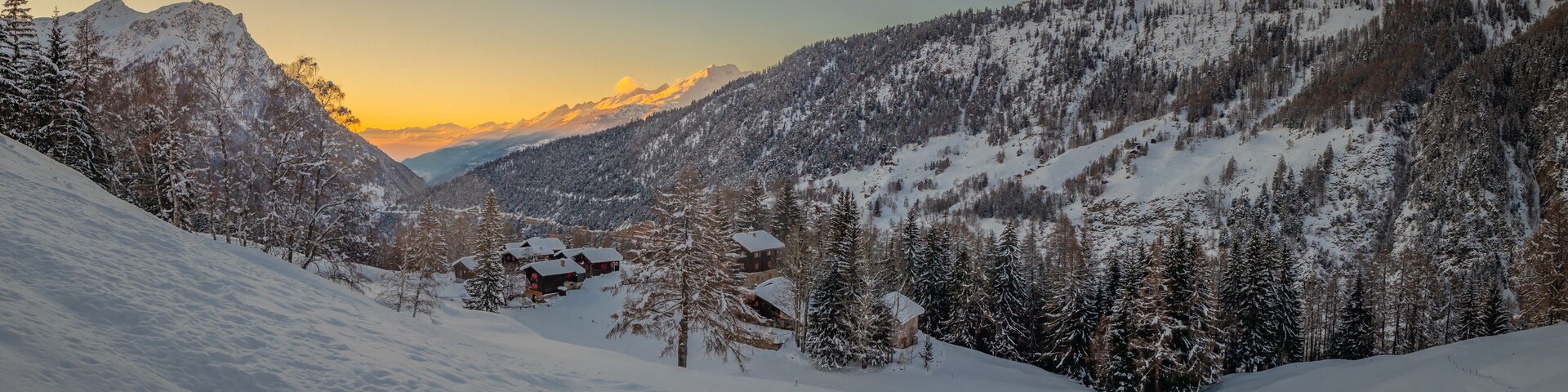 Paisagem montanhosa coberta de neve com um vale, casas e florestas densas.