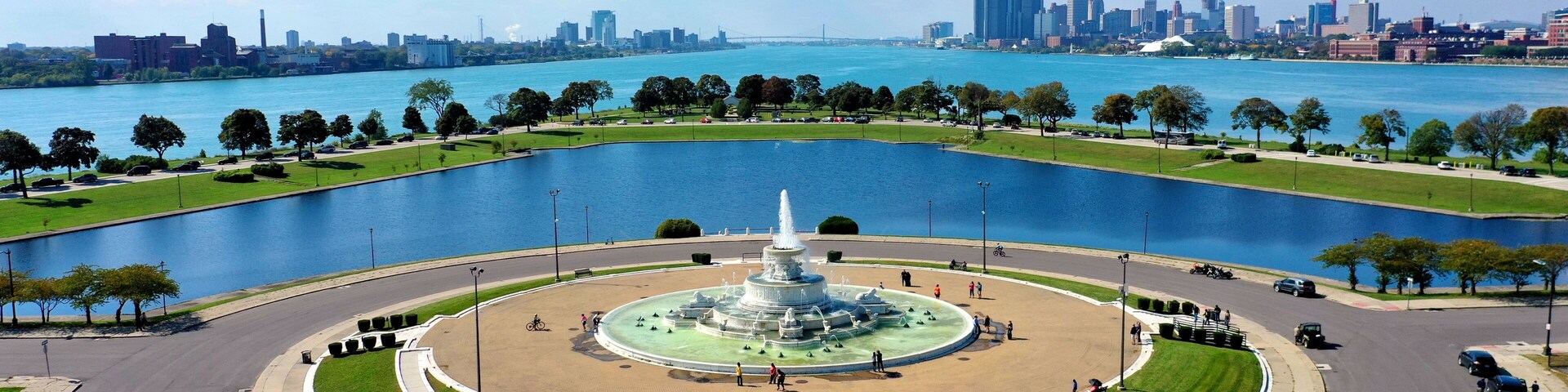 Paisagem urbana com uma grande extensão de água, uma fonte central e uma ponte ao longe.
