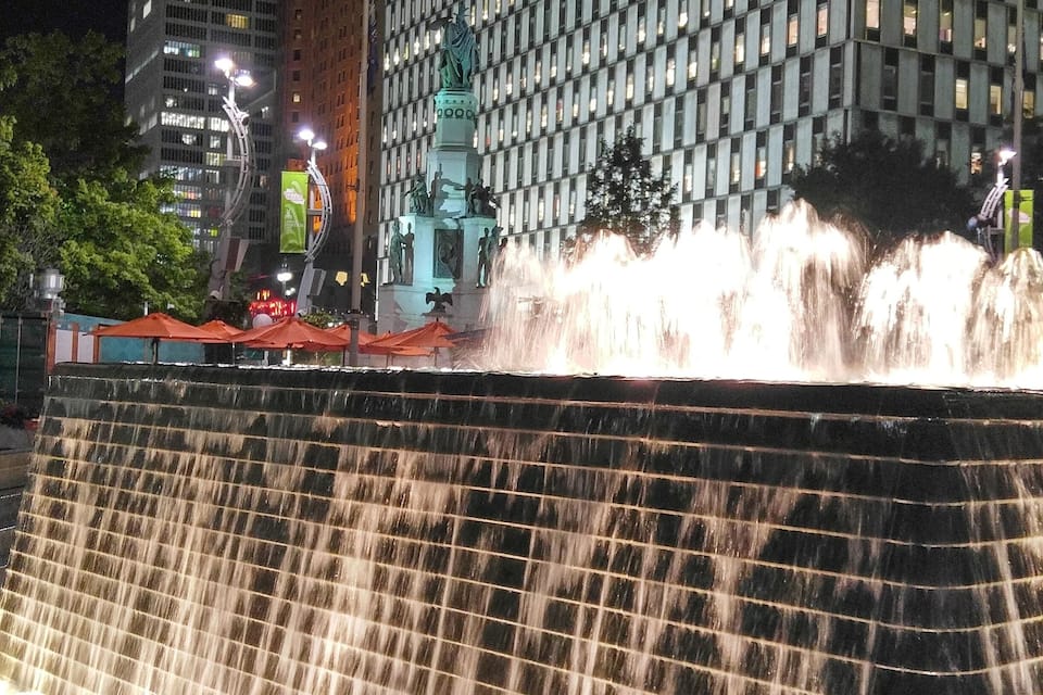 Uma cascata iluminada à noite, com uma estátua ao fundo.