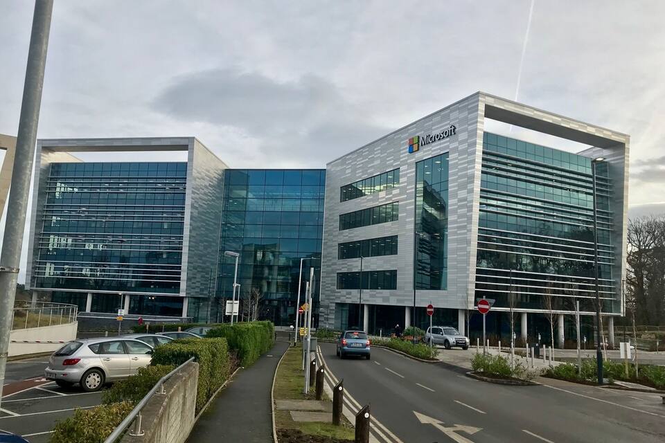 Um edifício moderno com o logotipo da Microsoft, rodeado por um parque de estacionamento e uma estrada com setas indicativas.