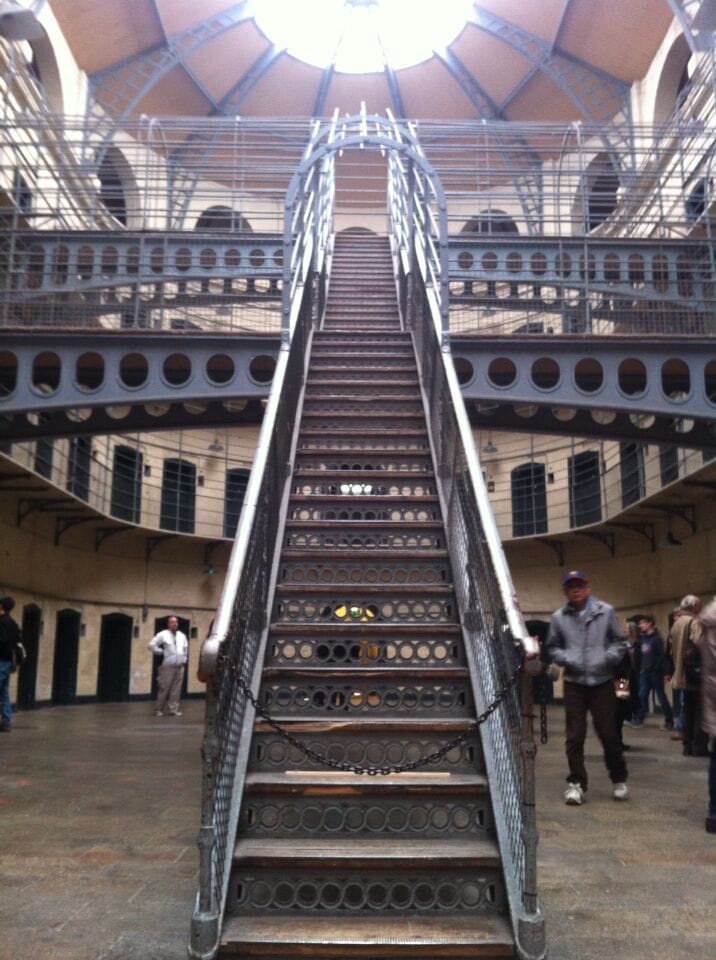 Uma escadaria imponente com grades de metal ricamente decoradas e padrões circulares, em um amplo átrio com pé-direito alto e janelas em arco.