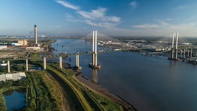 Uma grande ponte suspensa por cabos atravessa um rio largo, com edifícios industriais e uma área verde com lago à esquerda.