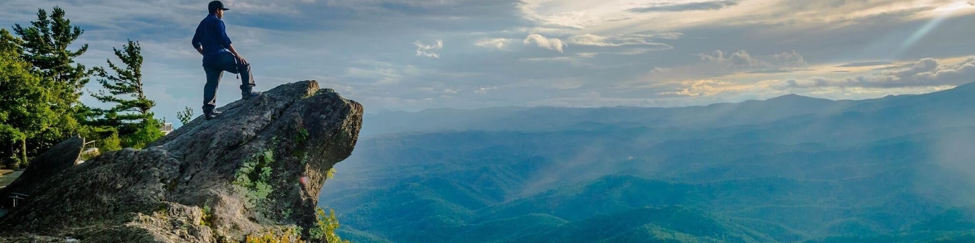 Uma pessoa está em um afloramento rochoso, com vista para uma paisagem montanhosa coberta por densa floresta.