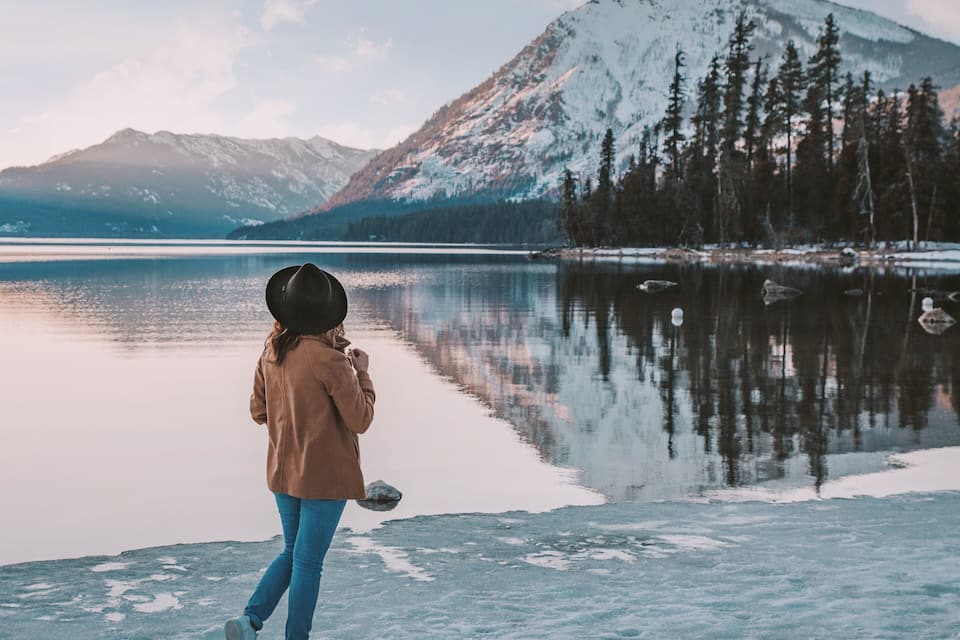 Uma pessoa parada à beira de um lago calmo, com montanhas cobertas de neve ao fundo.