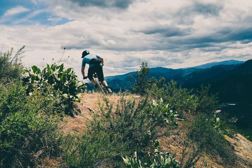 Um ciclista de montanha numa trilha, com vegetação exuberante e montanhas ao longe, sob um céu nublado.