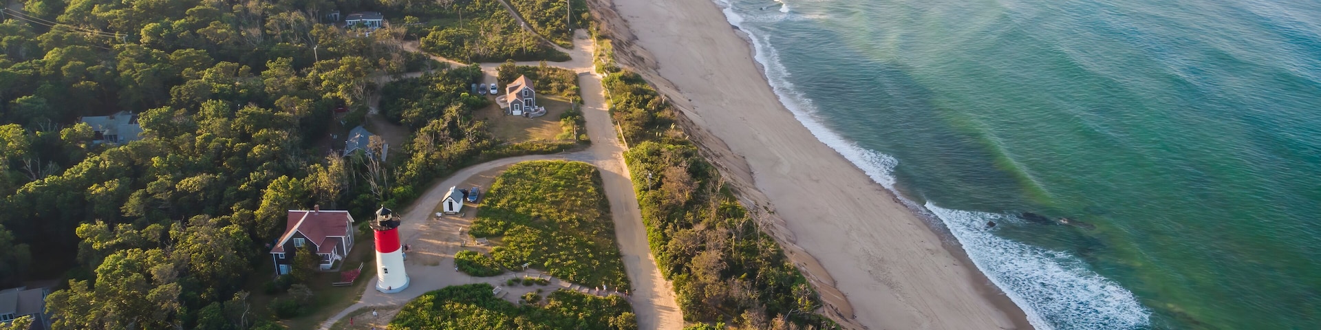 Vista aérea de uma área costeira com um farol, praia e floresta ao redor.