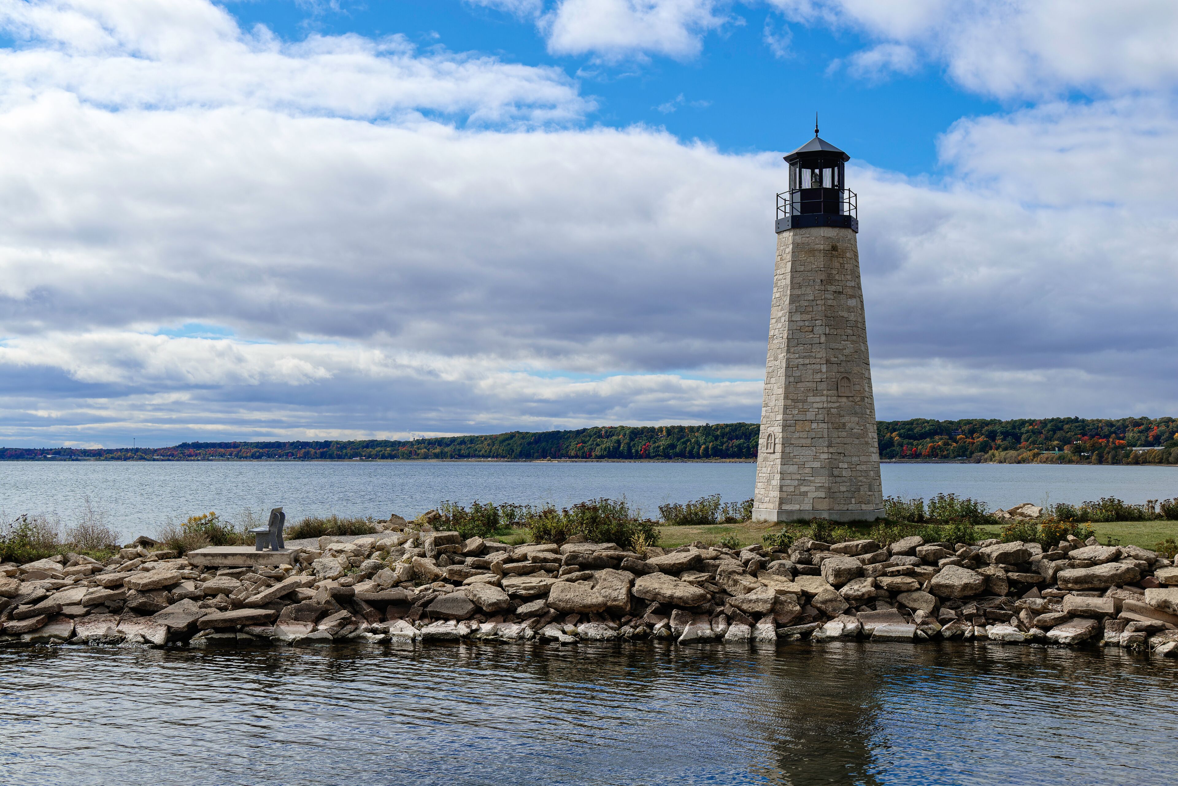 Um farol de pedra junto a uma costa rochosa, com um lago e colinas arborizadas ao fundo.