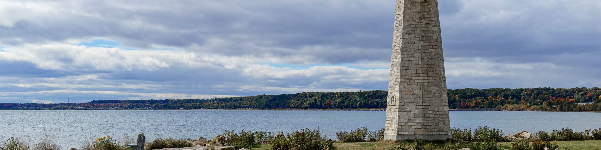 Um farol de pedra junto a uma costa rochosa, com um lago e colinas arborizadas ao fundo.