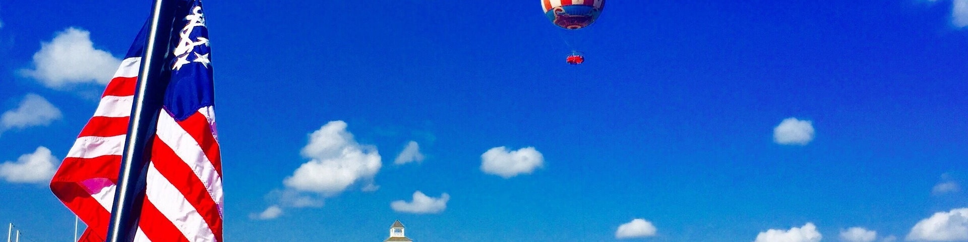Céu azul límpido com um balão de ar quente, uma bandeira e uma orla marítima com edifícios.