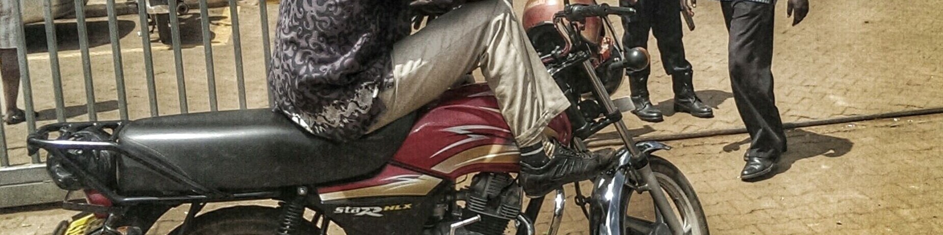 Um homem sentado numa motocicleta estacionada numa área pavimentada, com um guarda-corpo de metal ao fundo.