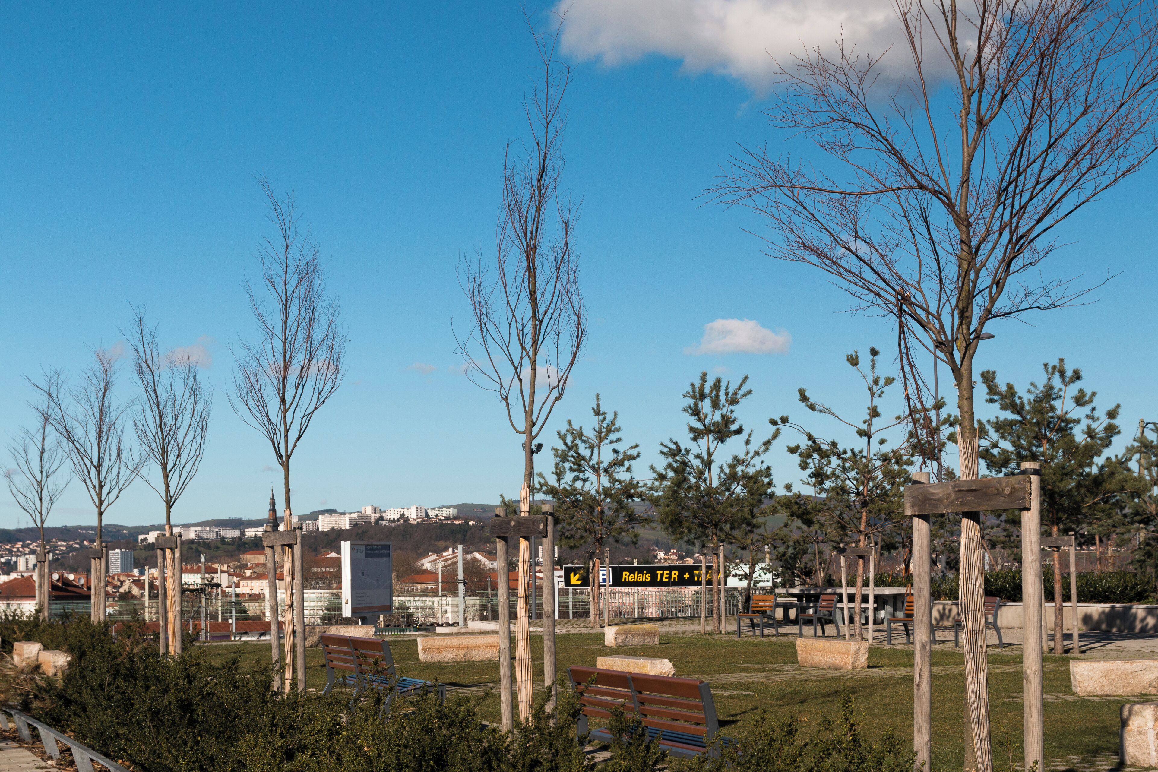 Um parque com bancos, árvores e uma placa indicando uma estação de trem.