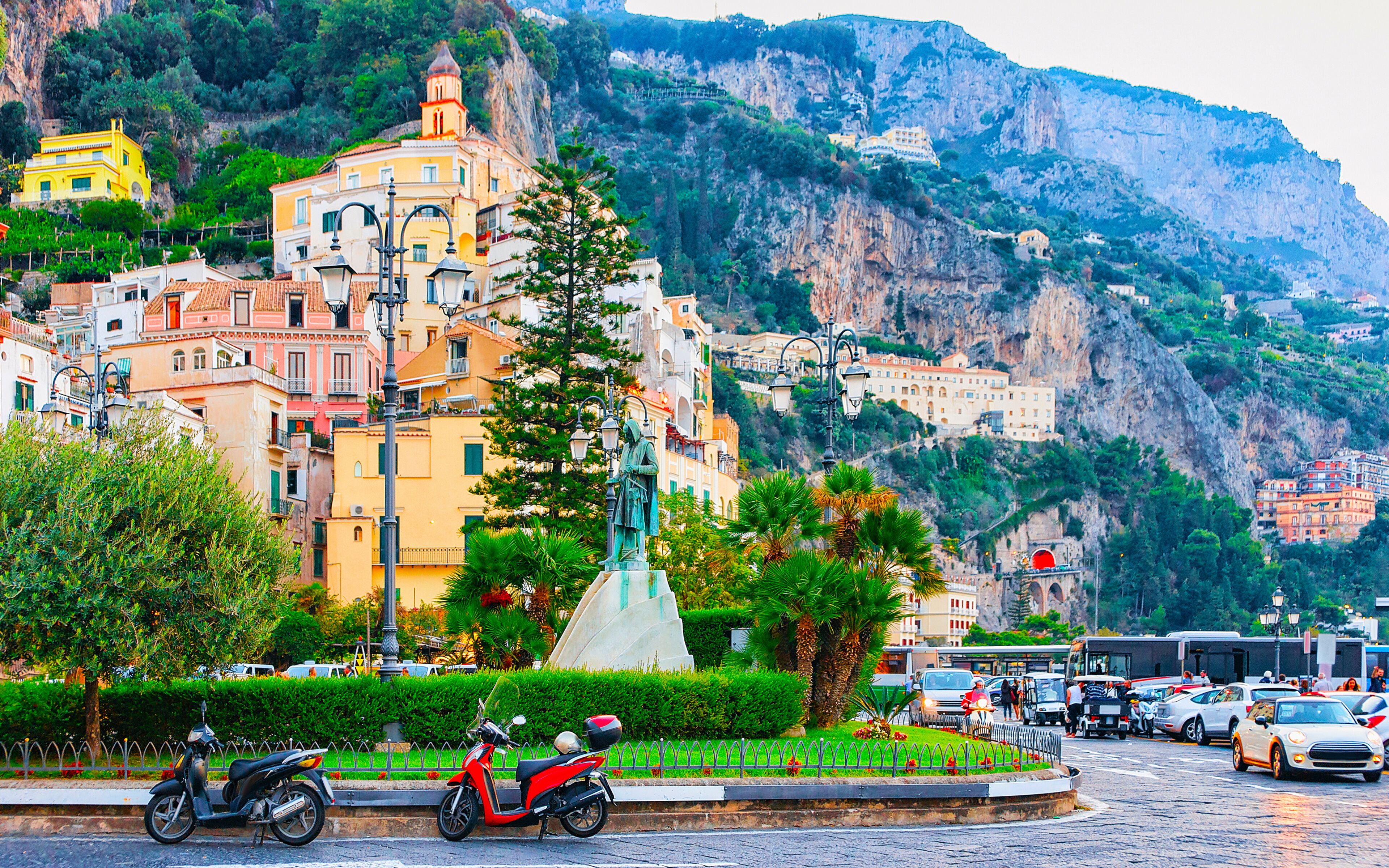 Uma vila costeira com edifícios coloridos, uma estátua e uma montanha ao fundo.