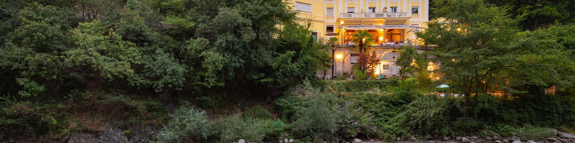 Um rio com pedras, cercado por árvores e um edifício amarelo com varandas.
