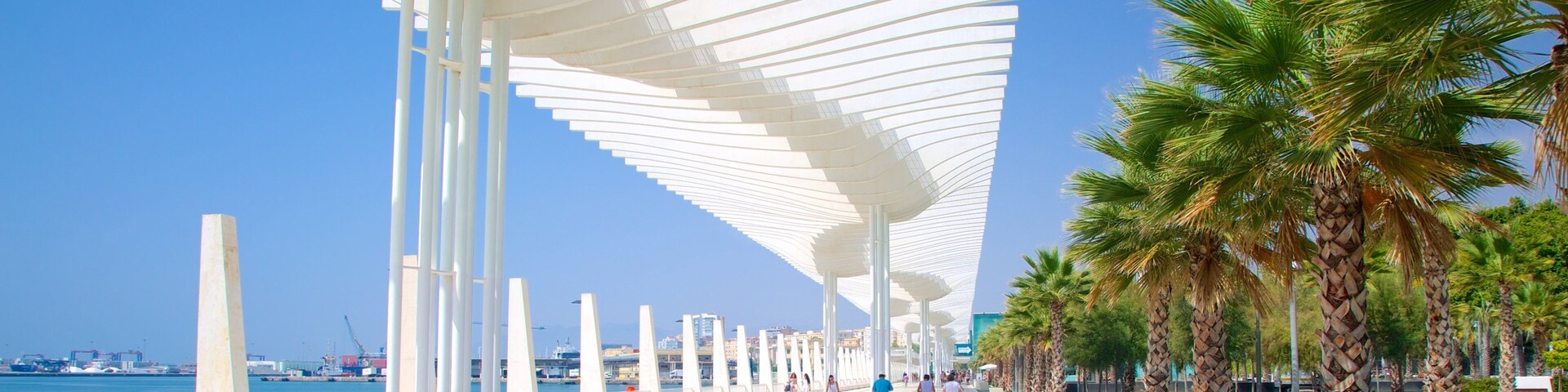 Um calçadão à beira-mar com uma moderna estrutura de toldo branco, palmeiras e um céu azul límpido.