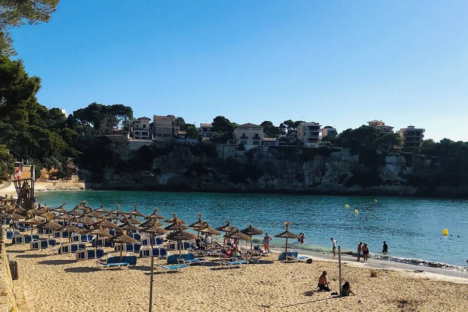 Uma praia com guarda-sóis e espreguiçadeiras, um penhasco rochoso com edifícios e água azul cristalina.