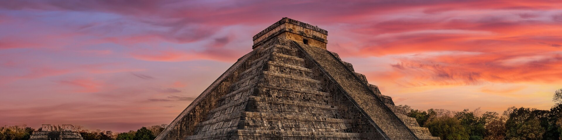 Uma grande estrutura piramidal com o topo plano, rodeada por uma área gramada e árvores, sob um céu colorido ao pôr do sol.