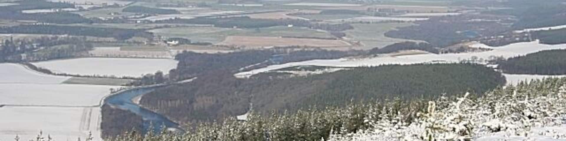 Paisagem nevada com rio, área arborizada e campos abertos.