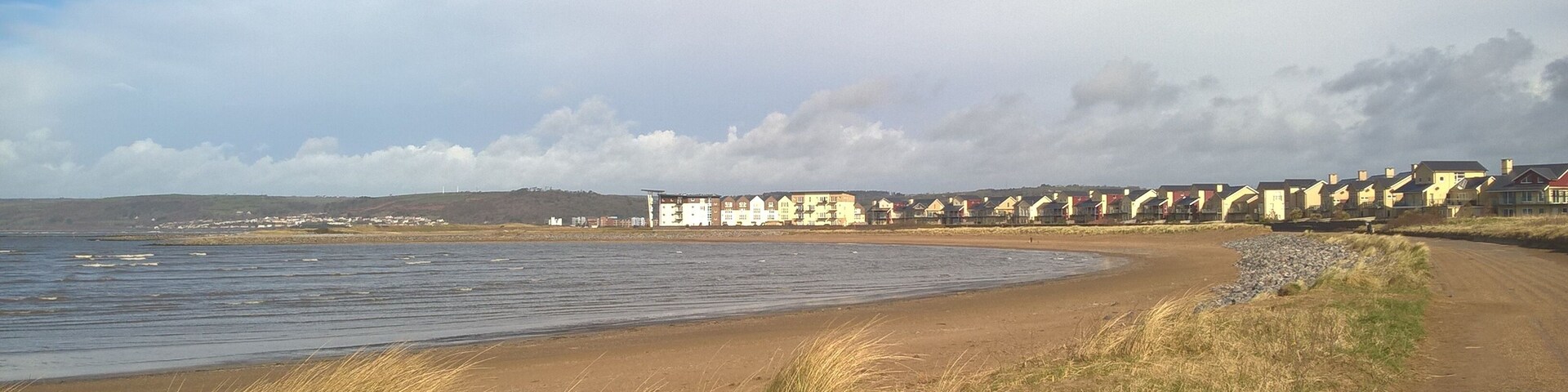 Paisagem costeira com praia de areia, dunas gramadas e fileira de casas à beira-mar.