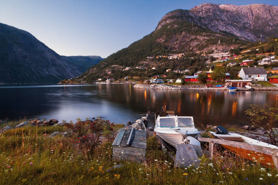Um lago tranquilo com barcos atracados num cais de madeira, rodeado por montanhas e casas.