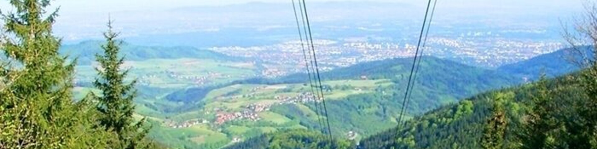 Um sistema de teleférico numa área montanhosa com florestas densas e uma vista distante de uma cidade.