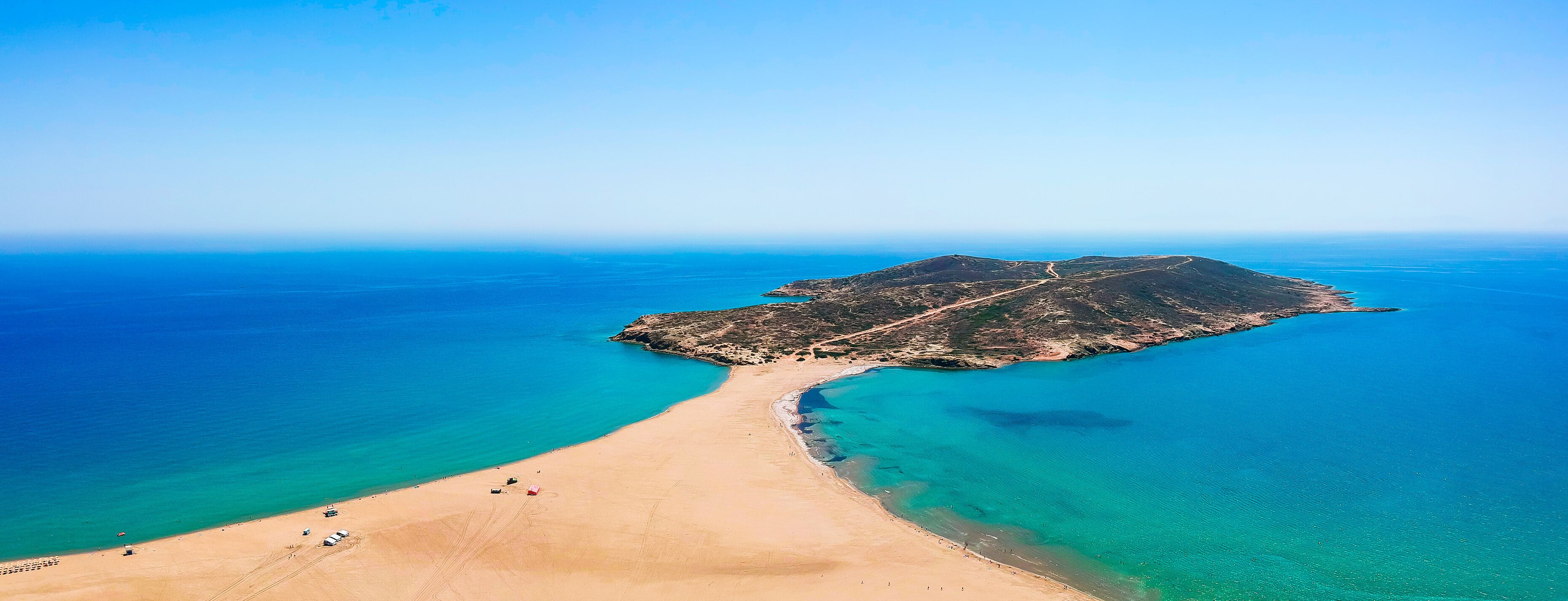 Vista aérea de uma área costeira com uma praia arenosa, águas turquesa cristalinas e uma pequena ilha rochosa.