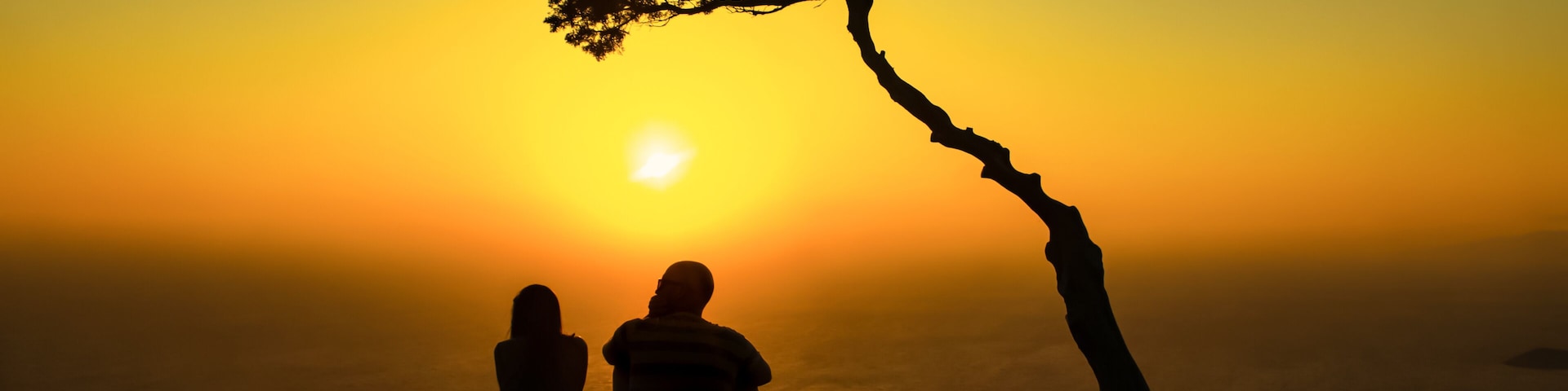Um pôr do sol com o perfil de uma árvore e duas pessoas sentadas em um afloramento rochoso.
