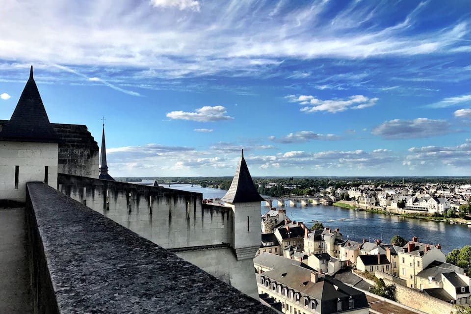 Uma cidade histórica com um rio, uma ponte e um castelo com torres cónicas.