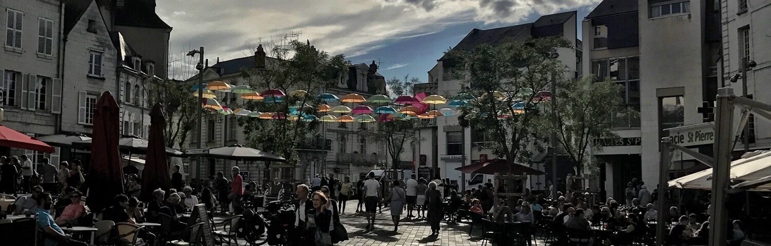 Uma praça de calçamento com mesas ao ar livre, rodeada por edifícios e um céu parcialmente nublado.