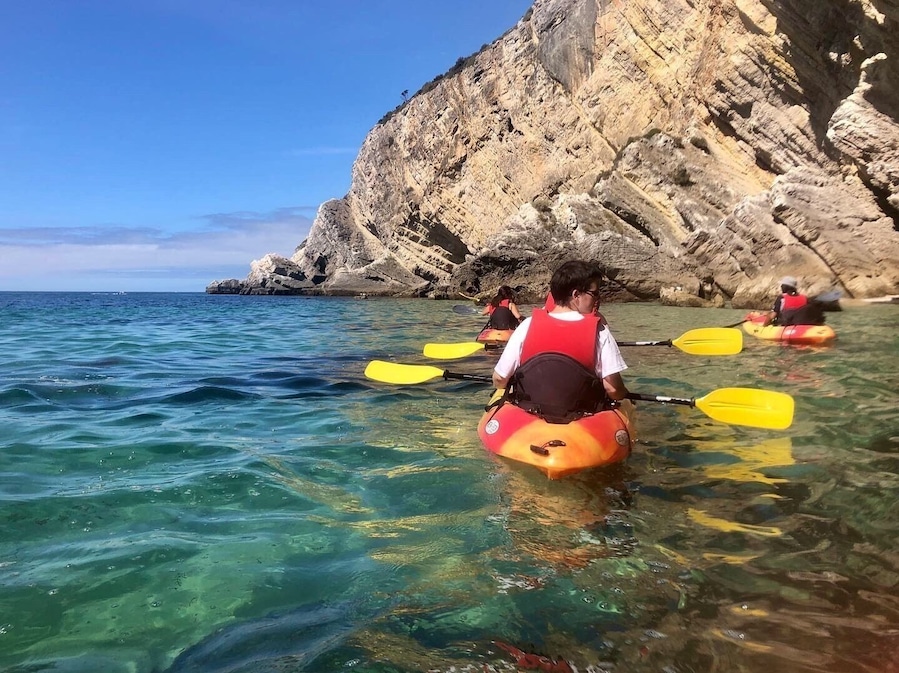 Caiaquistas em um mar calmo com penhascos rochosos ao fundo.