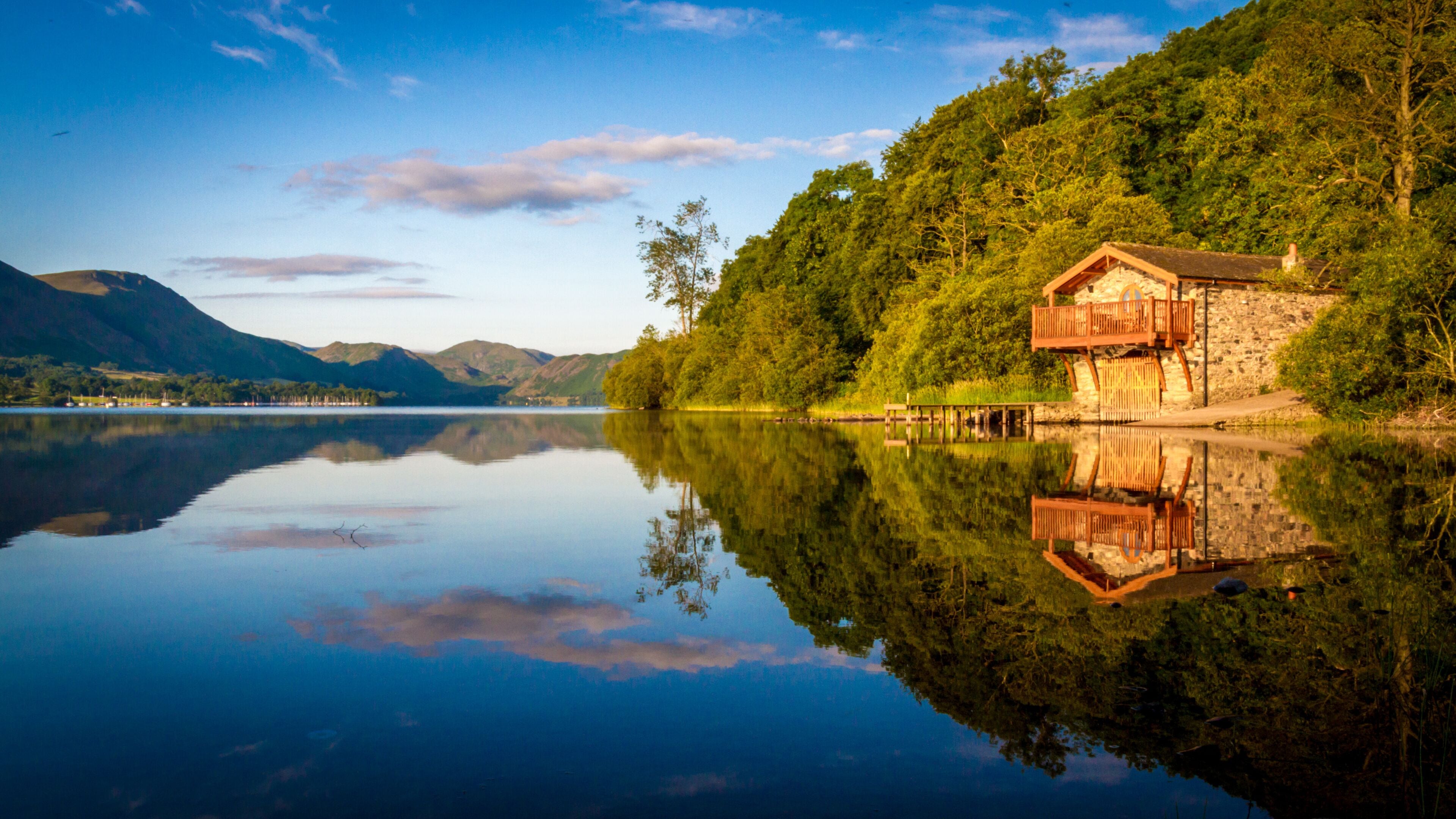 Um lago tranquilo com uma cabana de madeira refletida na água, rodeada por vegetação exuberante e montanhas ao fundo.