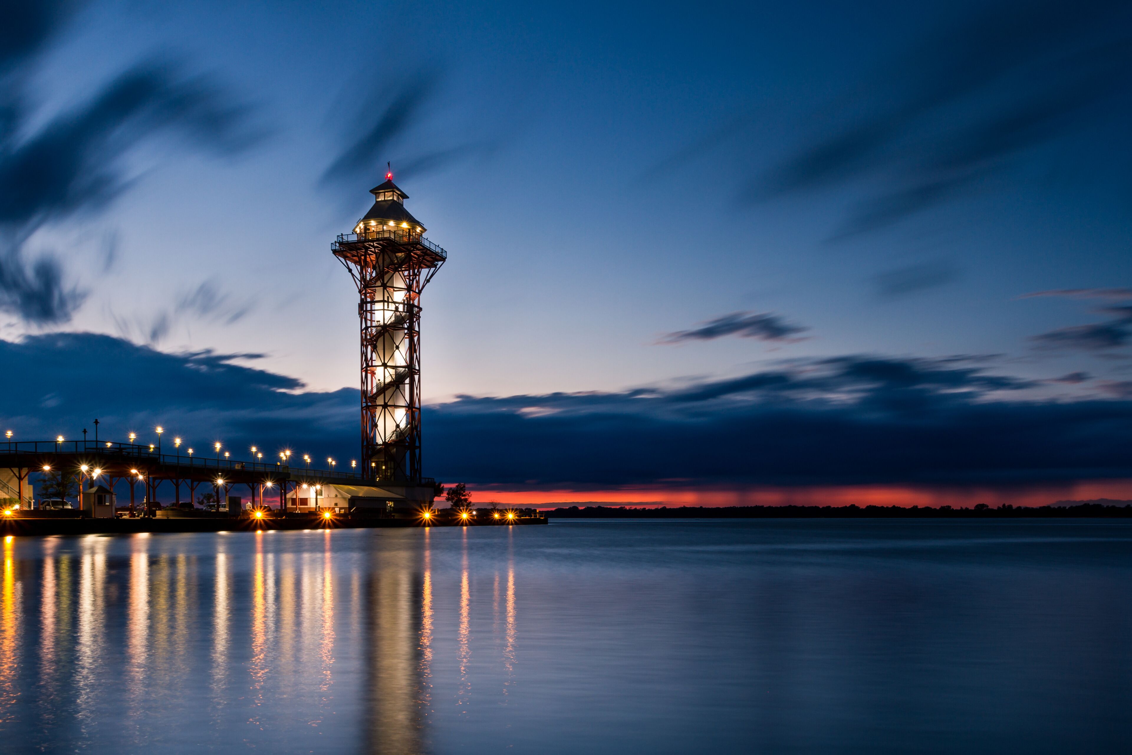 Um farol alto e iluminado, à beira de águas calmas ao entardecer.