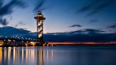 Um farol alto e iluminado, à beira de águas calmas ao entardecer.