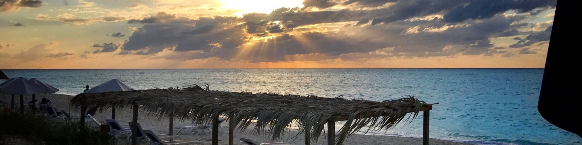Uma praia com cadeiras de descanso e uma estrutura de palha sob um céu ao pôr do sol.