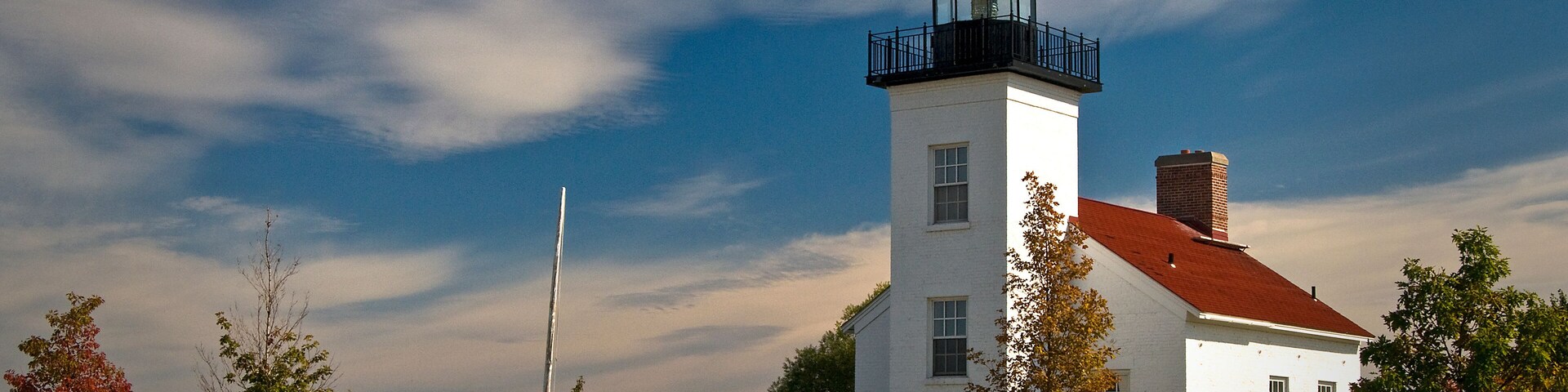 Um farol branco com teto vermelho e varanda preta, cercado por uma cerca branca e árvores.