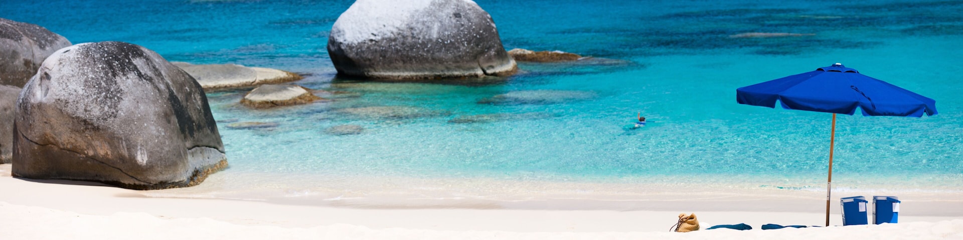 Uma praia com pedras grandes, água turquesa límpida e um guarda-sol azul.
