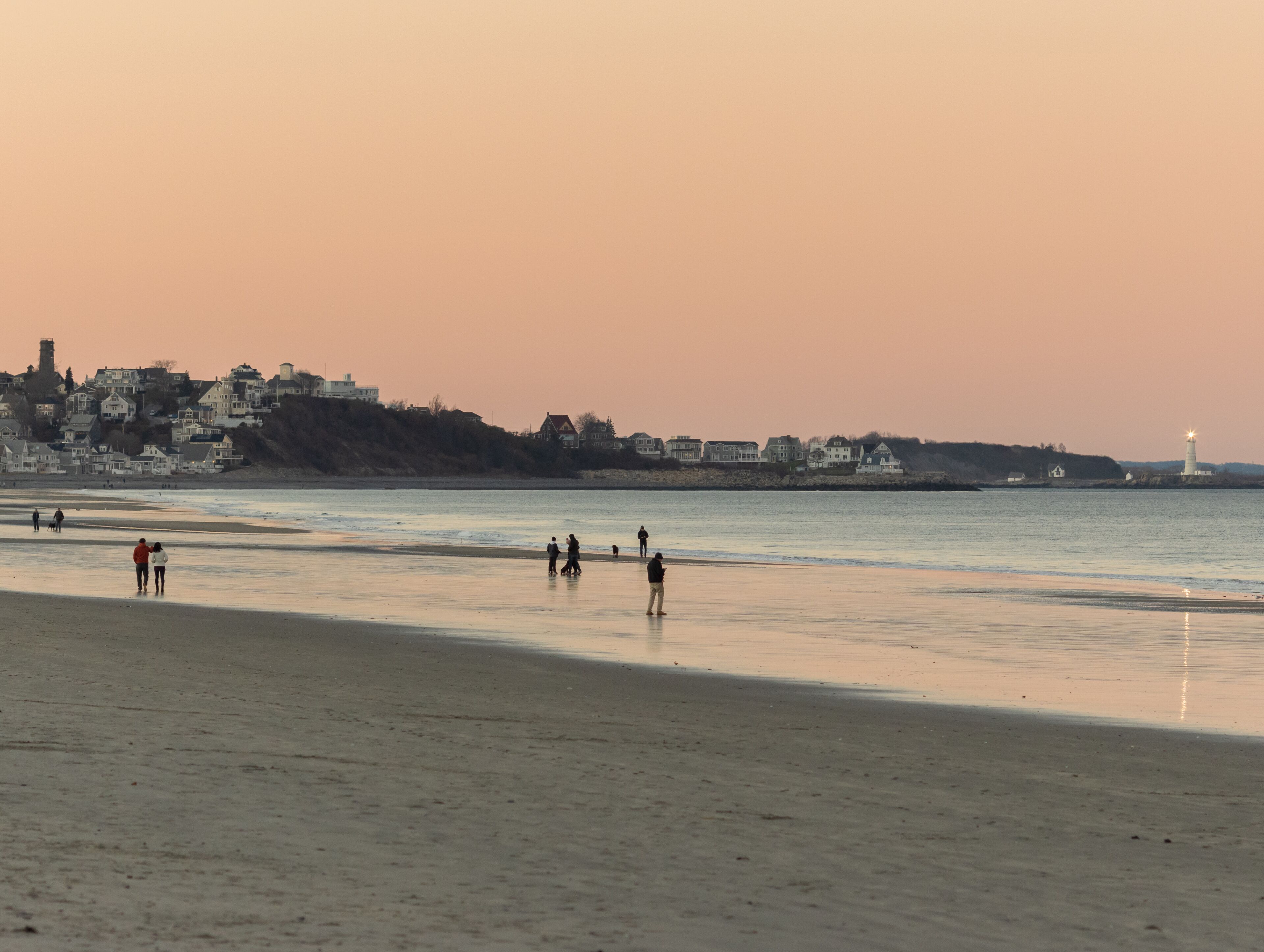 Uma praia com pessoas caminhando pela margem, um farol à distância e casas numa encosta.