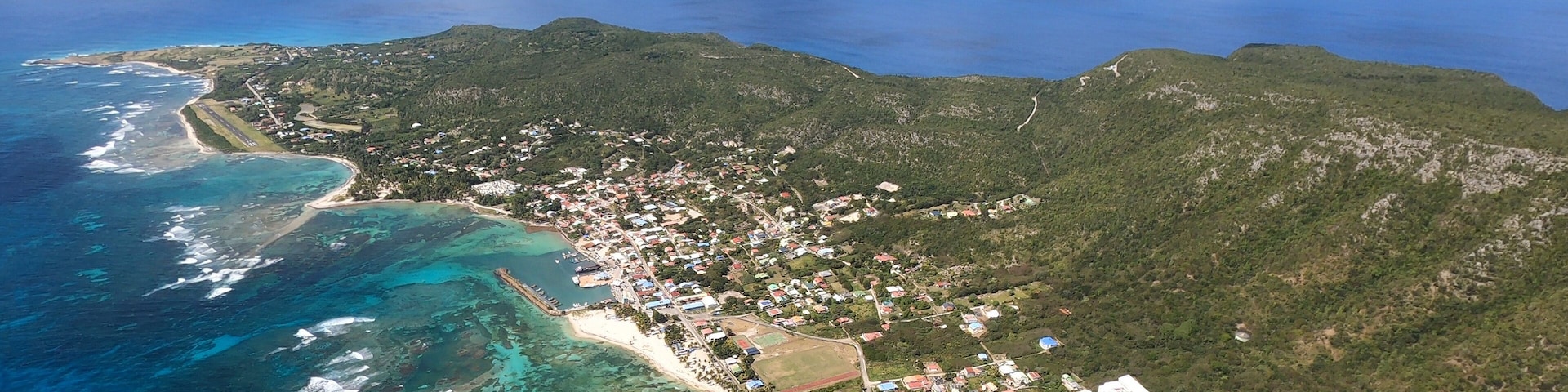 Vista aérea de uma cidade costeira com praia, águas azuis cristalinas e colinas verdejantes.