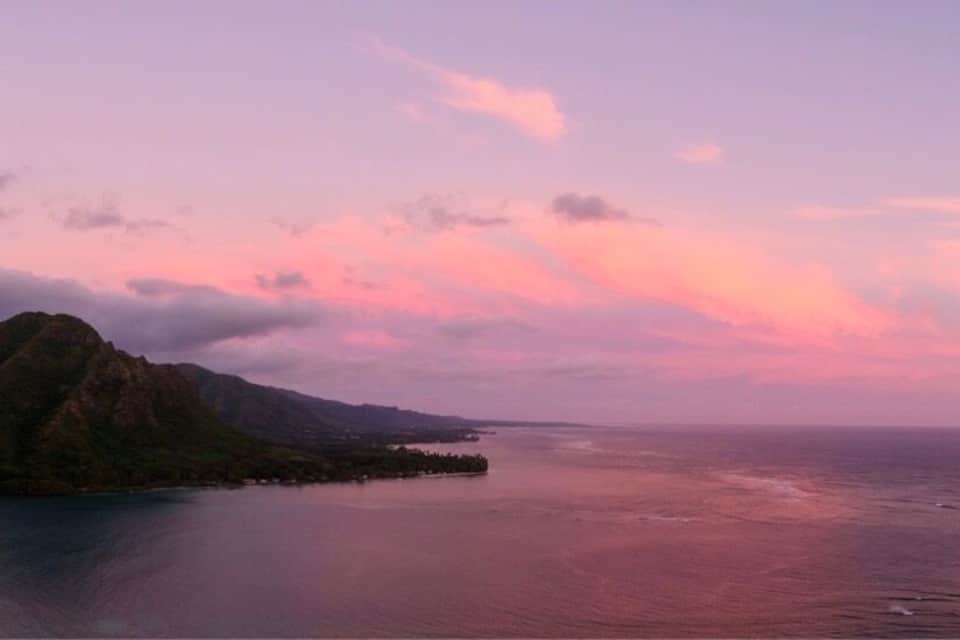 Paisagem costeira com uma cordilheira e um mar calmo sob um céu rosado e lilás.