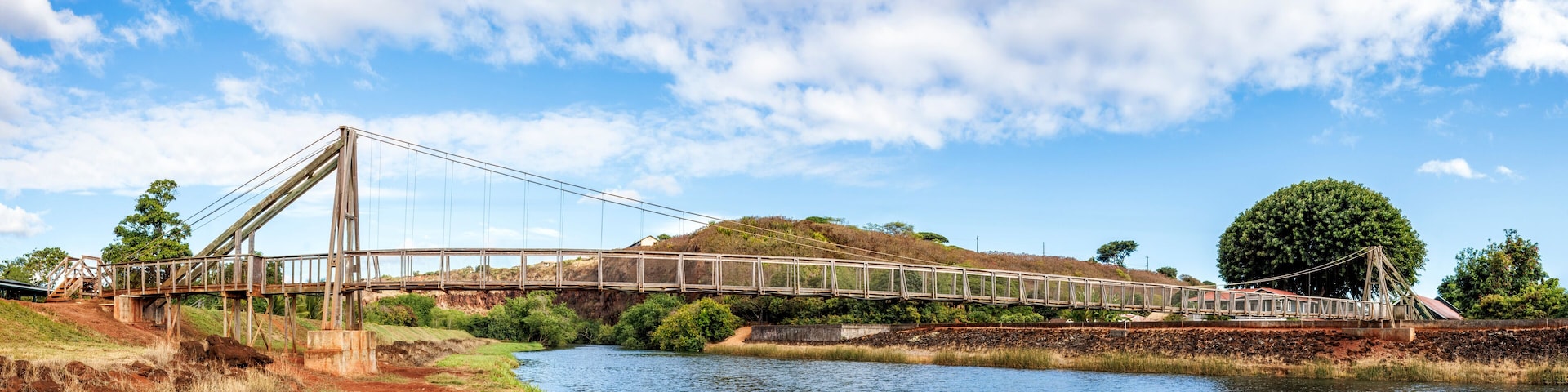 Uma ponte suspensa sobre um rio, com paisagem montanhosa e árvores ao fundo.