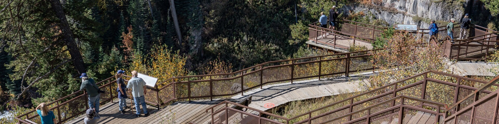 Um mirante de madeira com grades de metal oferece vista para um vale arborizado e uma cachoeira.