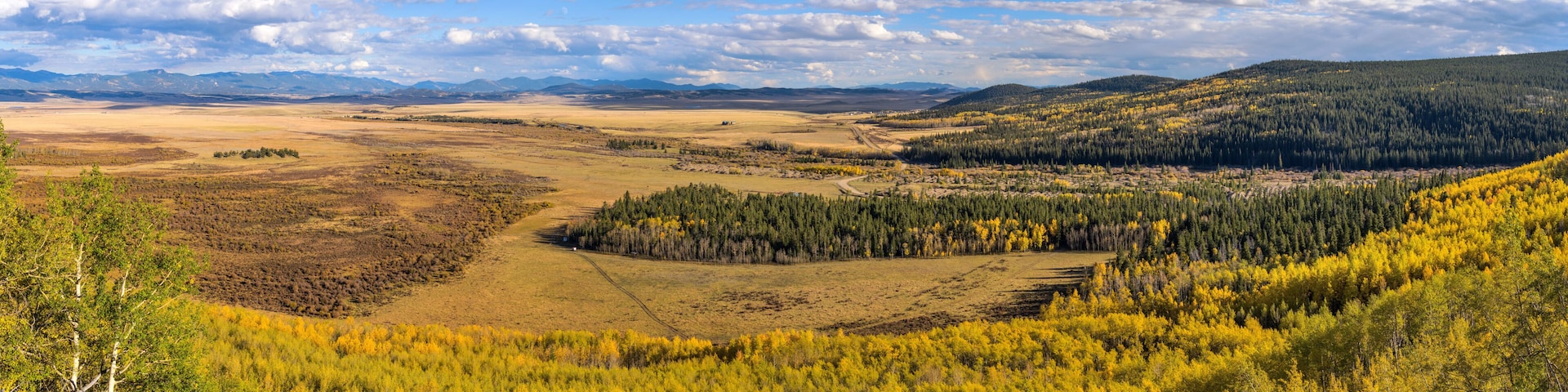 Extensa paisagem com áreas arborizadas e campos abertos, uma estrada sinuosa e montanhas distantes sob um céu parcialmente nublado.