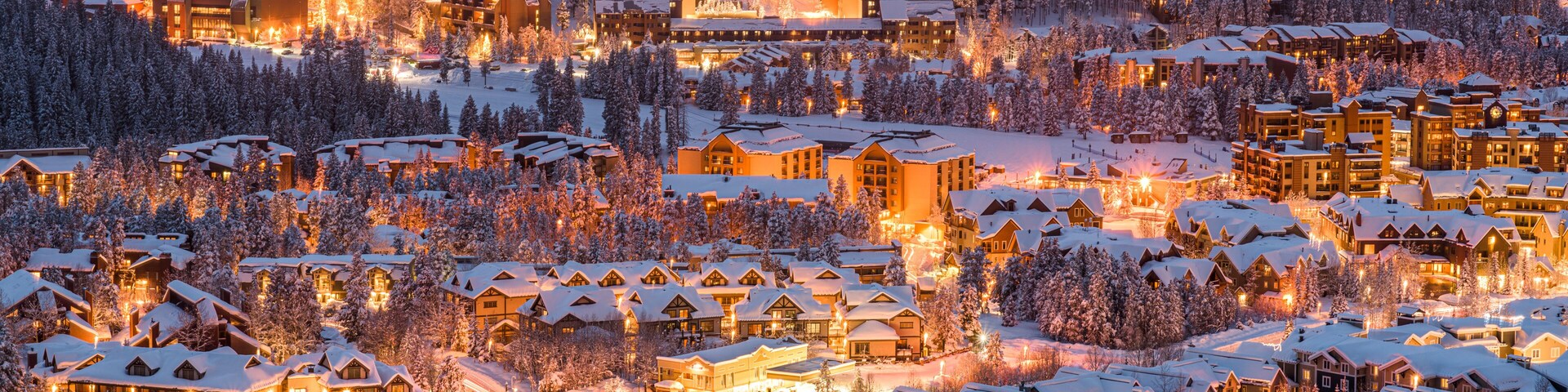 Cidade turística coberta de neve ao entardecer, com edifícios iluminados e uma estrada sinuosa.