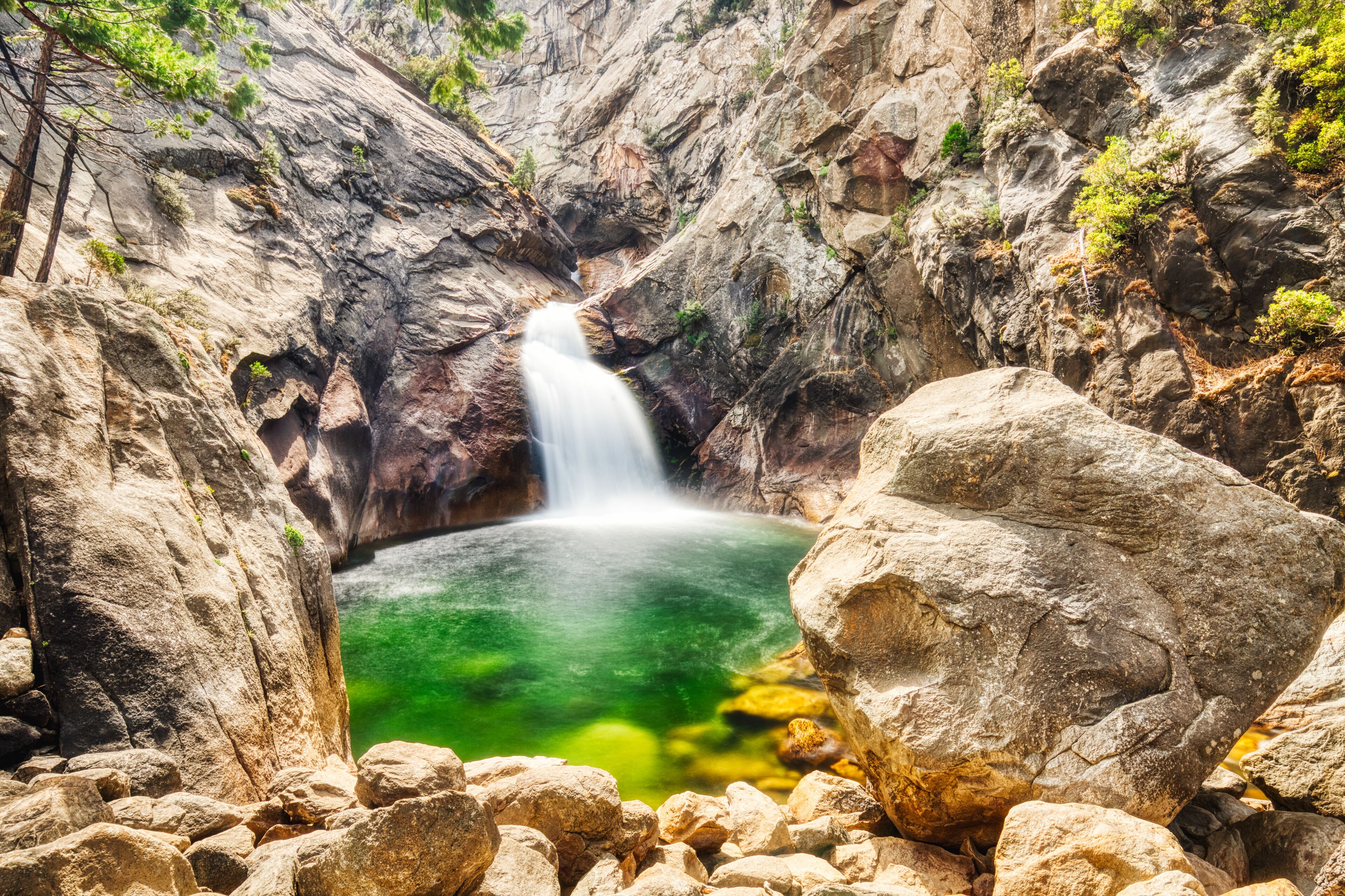 Uma cascata deságua em uma piscina verde e límpida, cercada por terreno rochoso e vegetação rasteira.