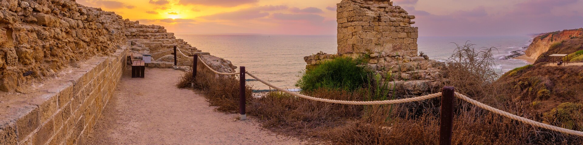 Um caminho costeiro que leva a ruínas antigas, com o pôr do sol ao fundo.