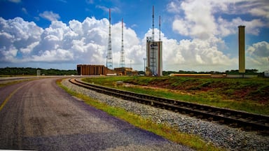 Trilhos de ferrovia curvando-se à direita, com uma torre de lançamento e uma chaminé alta ao fundo.