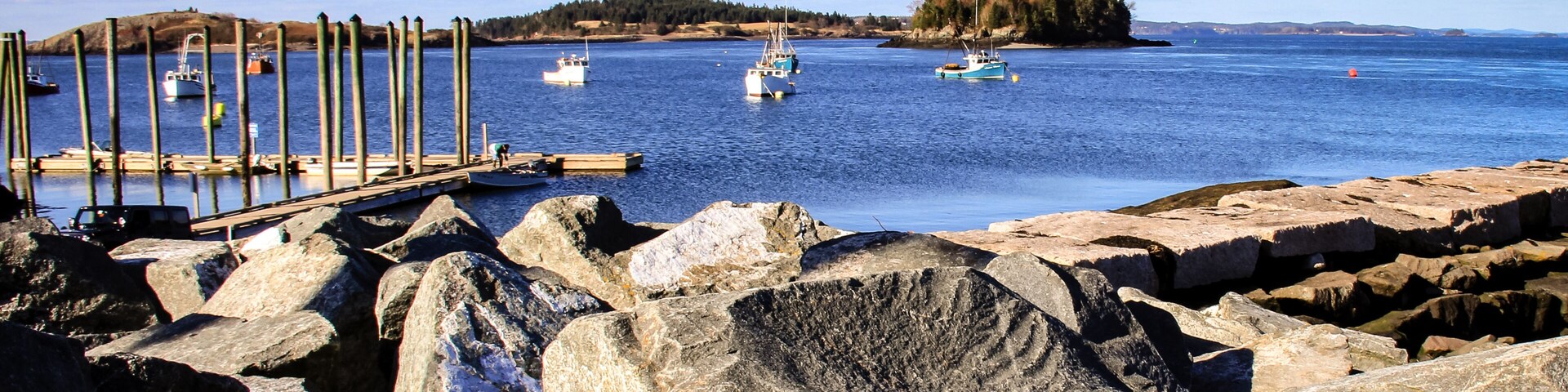Uma costa rochosa com barcos em um mar calmo, um cais de madeira e uma ilha distante coberta de árvores.