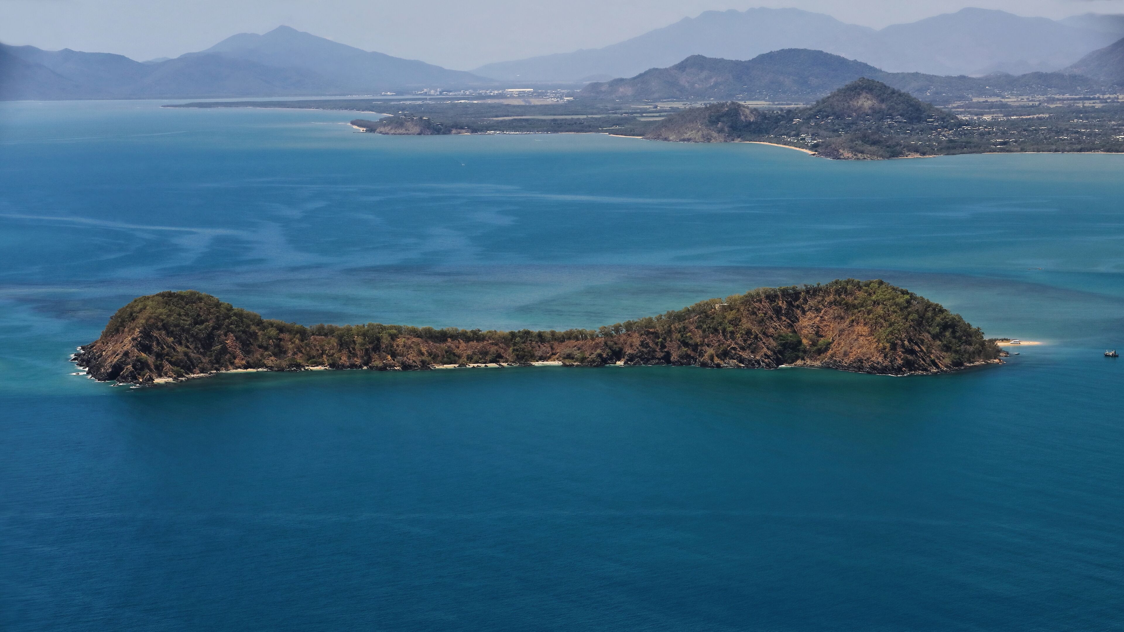 Vista aérea de uma ilha grande cercada por águas azuis cristalinas, com montanhas ao fundo.